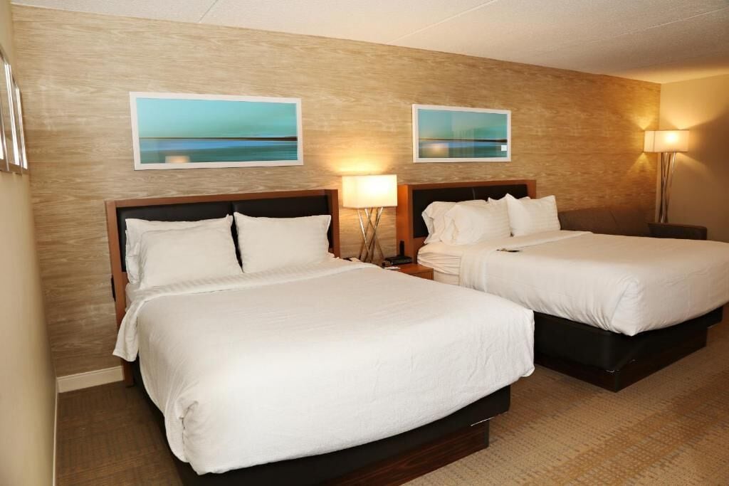 Quarto de hotel com duas camas, cabeceira de madeira, mesa de cabeceira com abajur e dois quadros na parede.