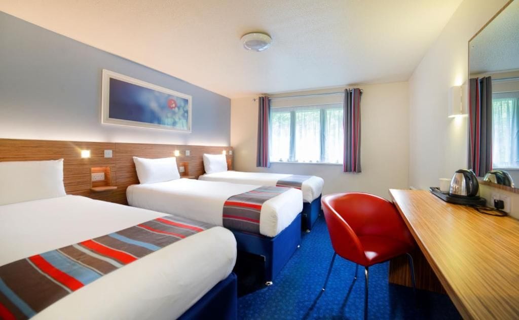 Quarto de hotel com duas camas, uma escrivaninha, uma cadeira e uma janela grande com cortinas.