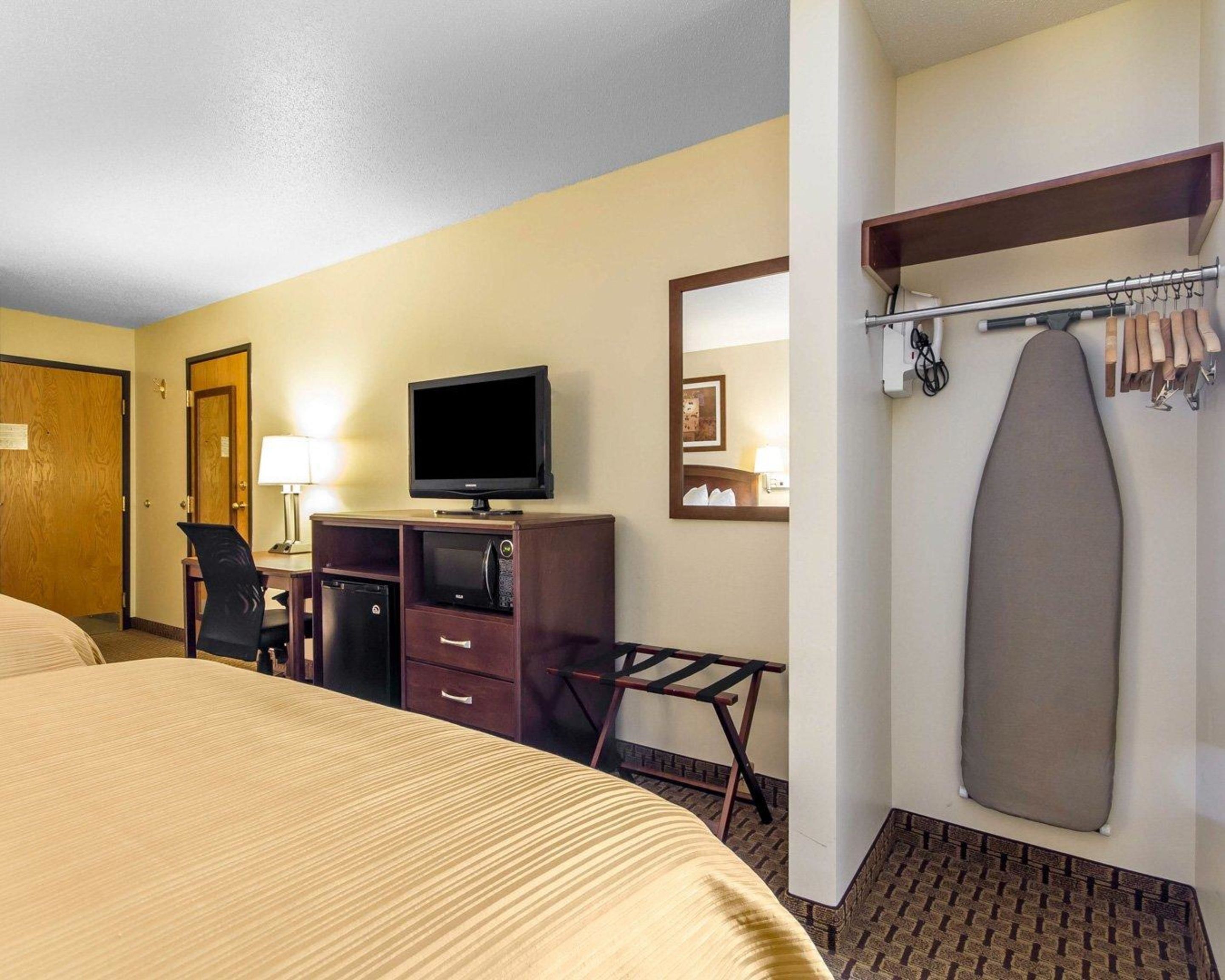 Quarto de hotel com cama, escrivaninha com computador, televisão, micro-ondas e tábua de passar roupa.