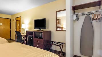 Quarto de hotel com cama, escrivaninha com computador, televisão, micro-ondas e tábua de passar roupa.