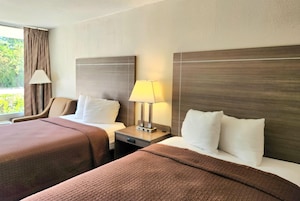 Quarto de hotel com duas camas, uma mesa de cabeceira com abajur, uma cadeira e uma janela com cortinas.
