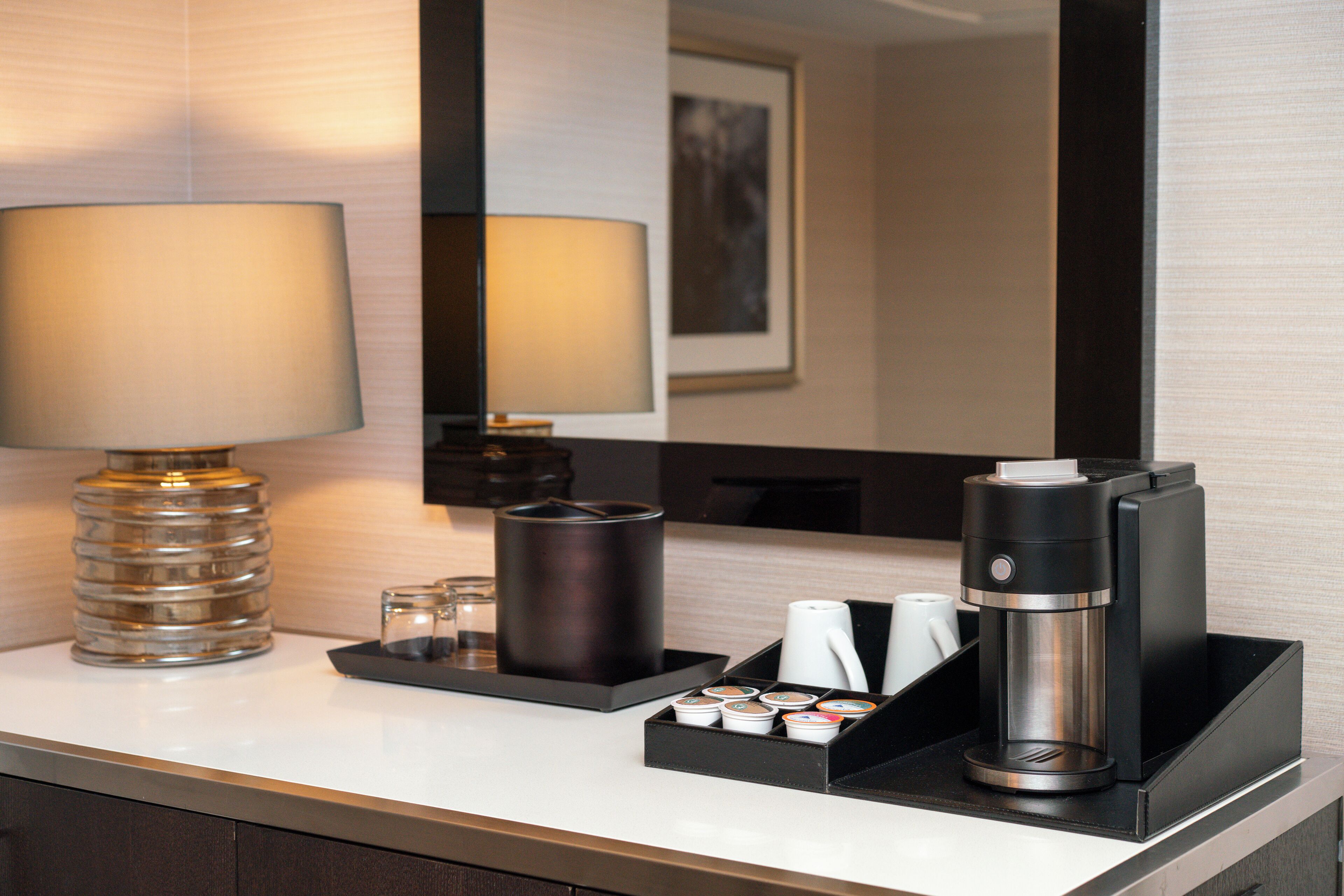 Balcão de quarto de hotel com cafeteira, luminária e bandeja com cápsulas de café.