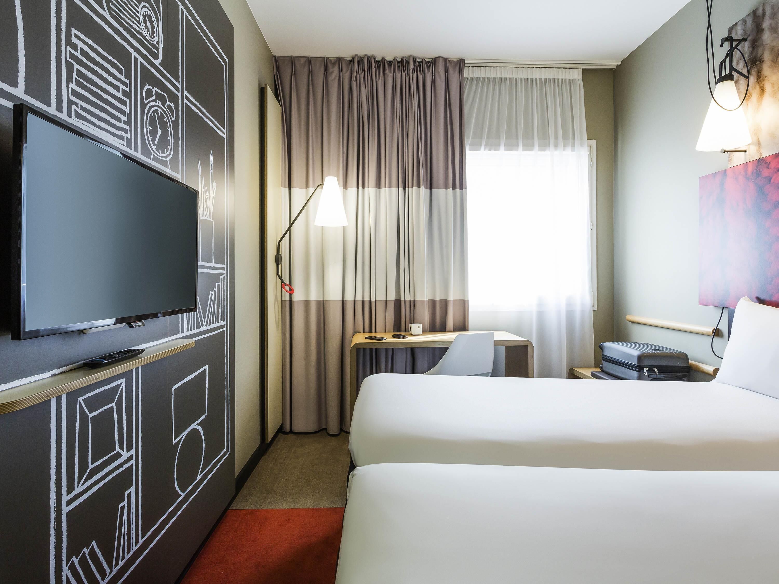 Quarto de hotel com uma cama, uma escrivaninha com cadeira, televisão e uma janela com cortinas.