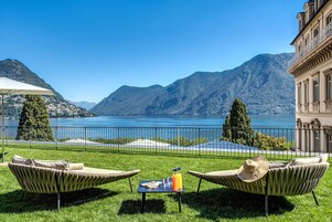 Terraço com duas cadeiras estofadas, uma mesa com bebida e vista para um lago com montanhas ao fundo.