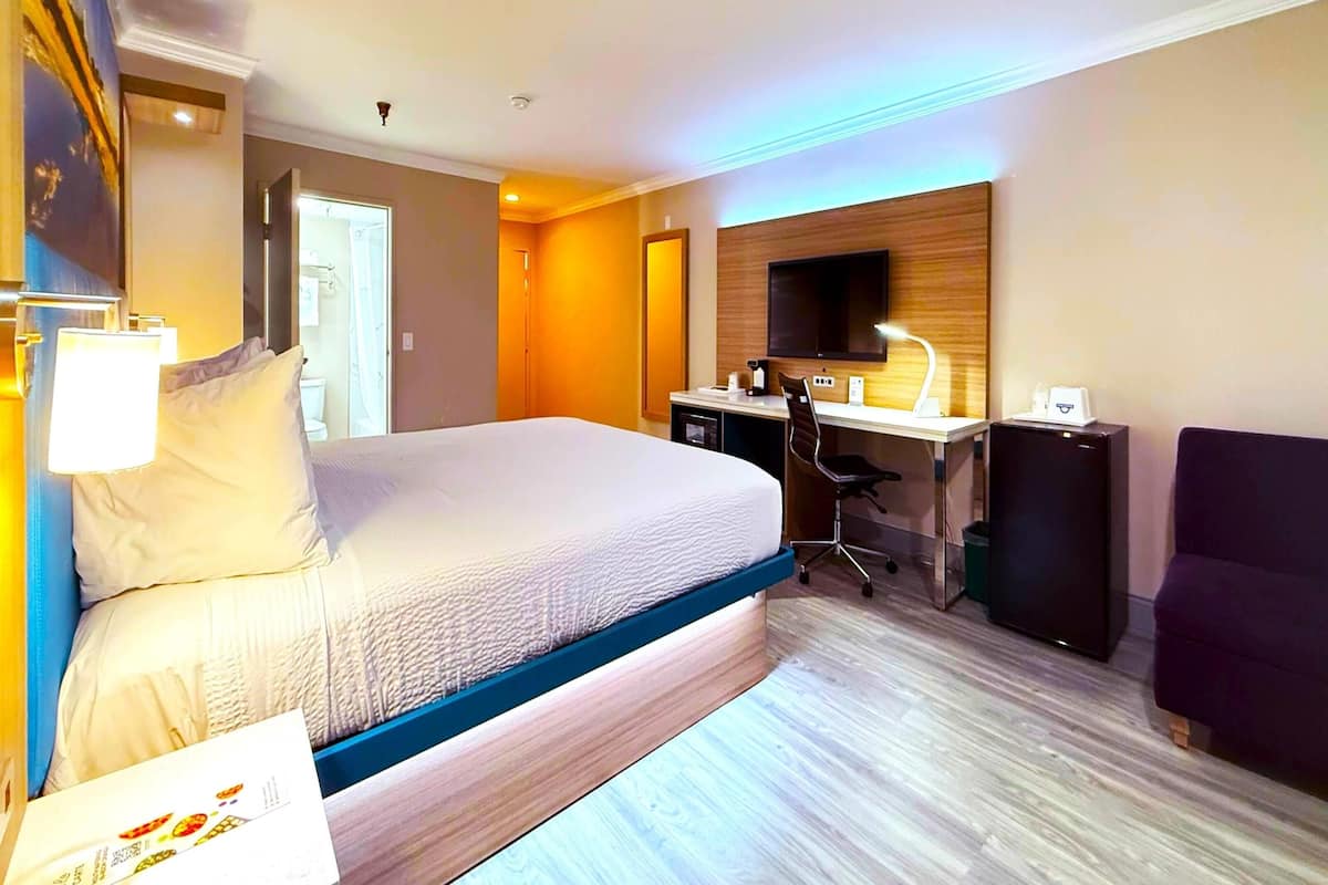 Quarto de hotel com uma cama grande, uma escrivaninha com computador, televisão e uma poltrona roxa.