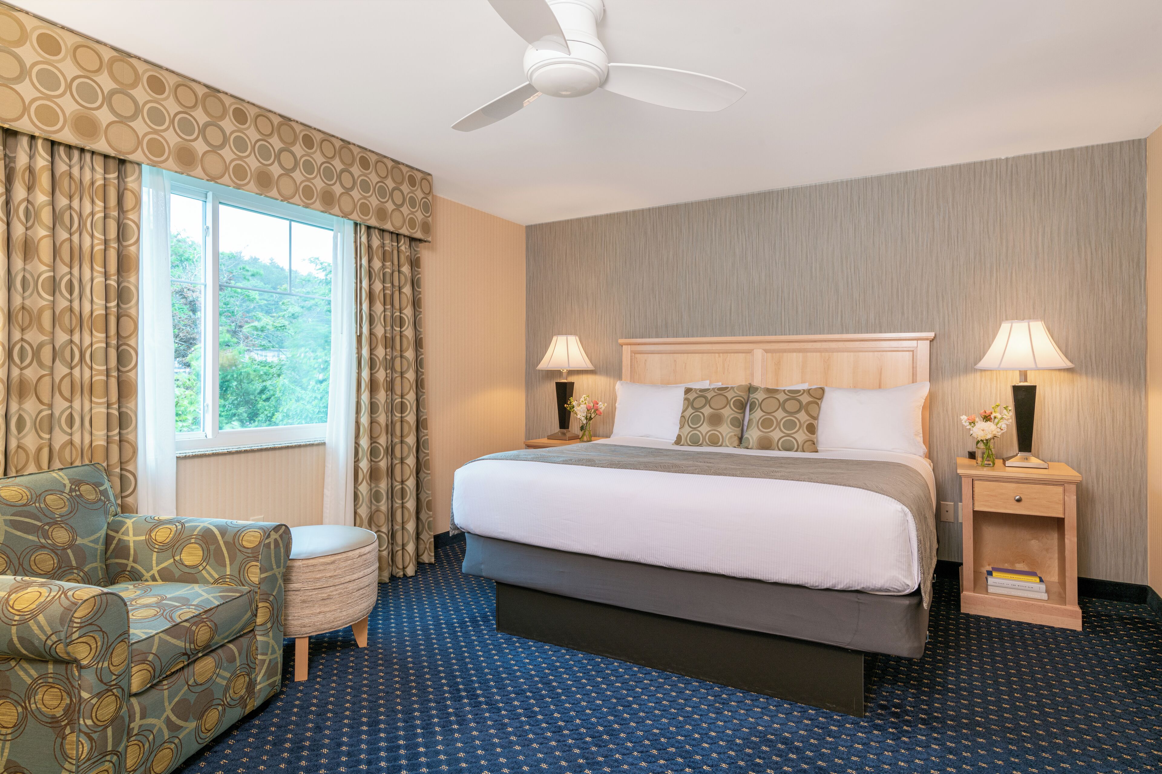 Quarto de hotel com cama, duas mesas de cabeceira com abajures, uma cadeira, janela com cortinas e um ventilador de teto.
