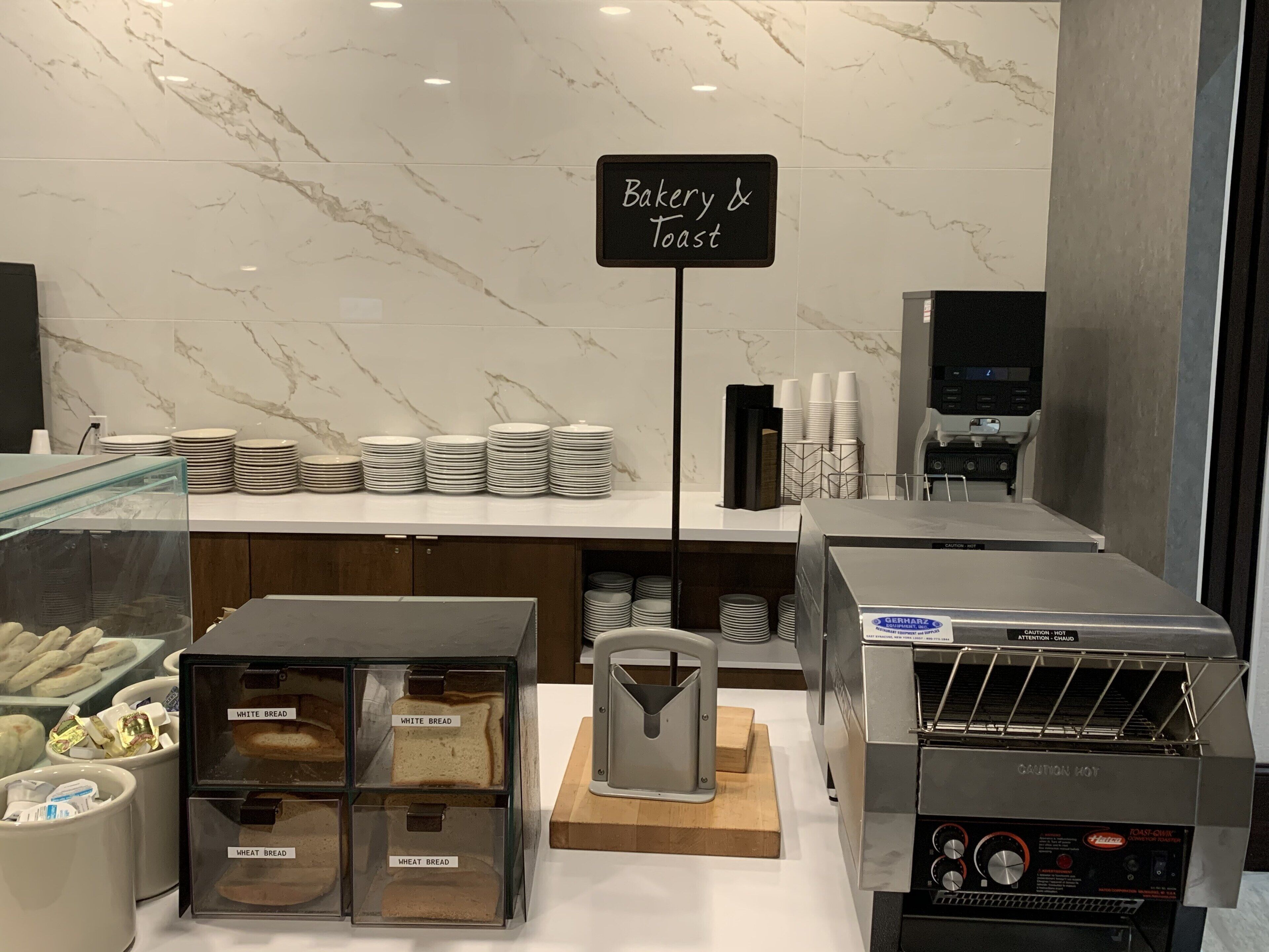 Balcão de pastelaria com pilhas de pratos, pães expostos e um aviso.