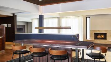 Uma área de bar moderna com um longo balcão de madeira, banquetas pretas e uma lareira ao fundo.