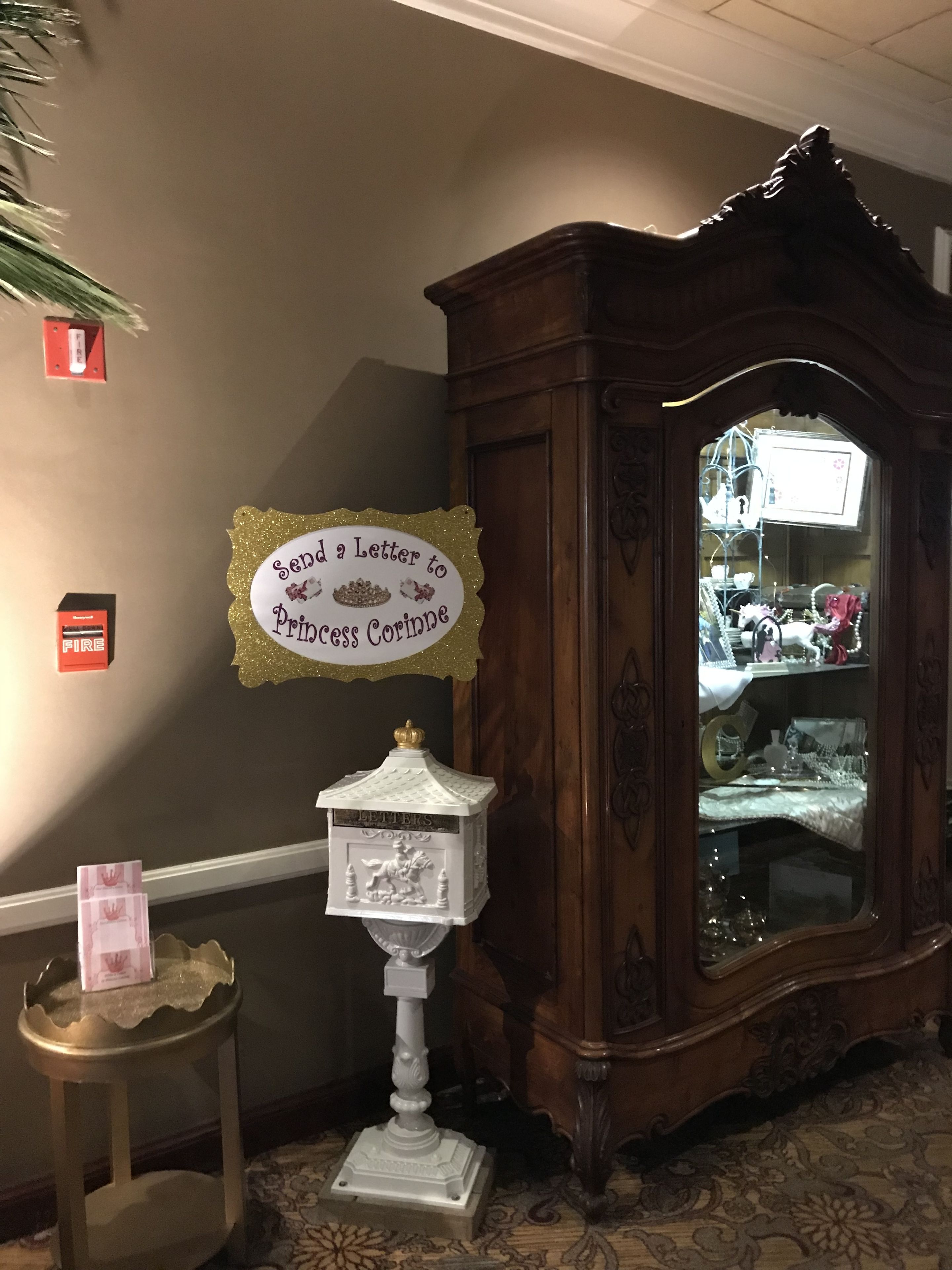 Um armário de madeira estilo vintage com portas de vidro, um pequeno pedestal branco com topo decorativo e uma placa na parede.