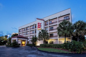 Um hotel com vários andares, um letreiro chamativo da 'Red Roof', estacionamento e áreas verdes ao redor.