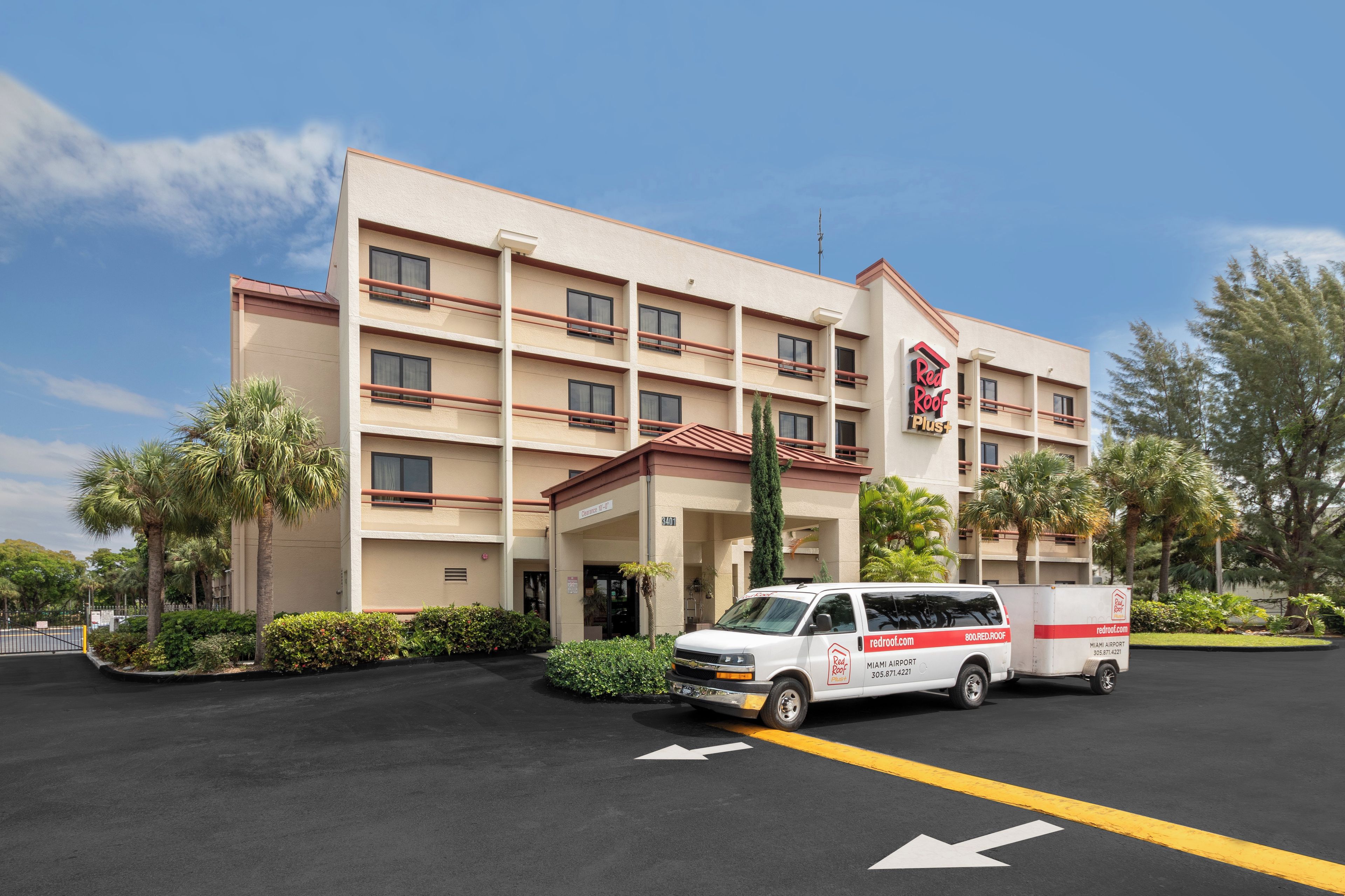 Um hotel de vários andares com placa de telhado vermelha, um estacionamento com uma van branca e palmeiras.