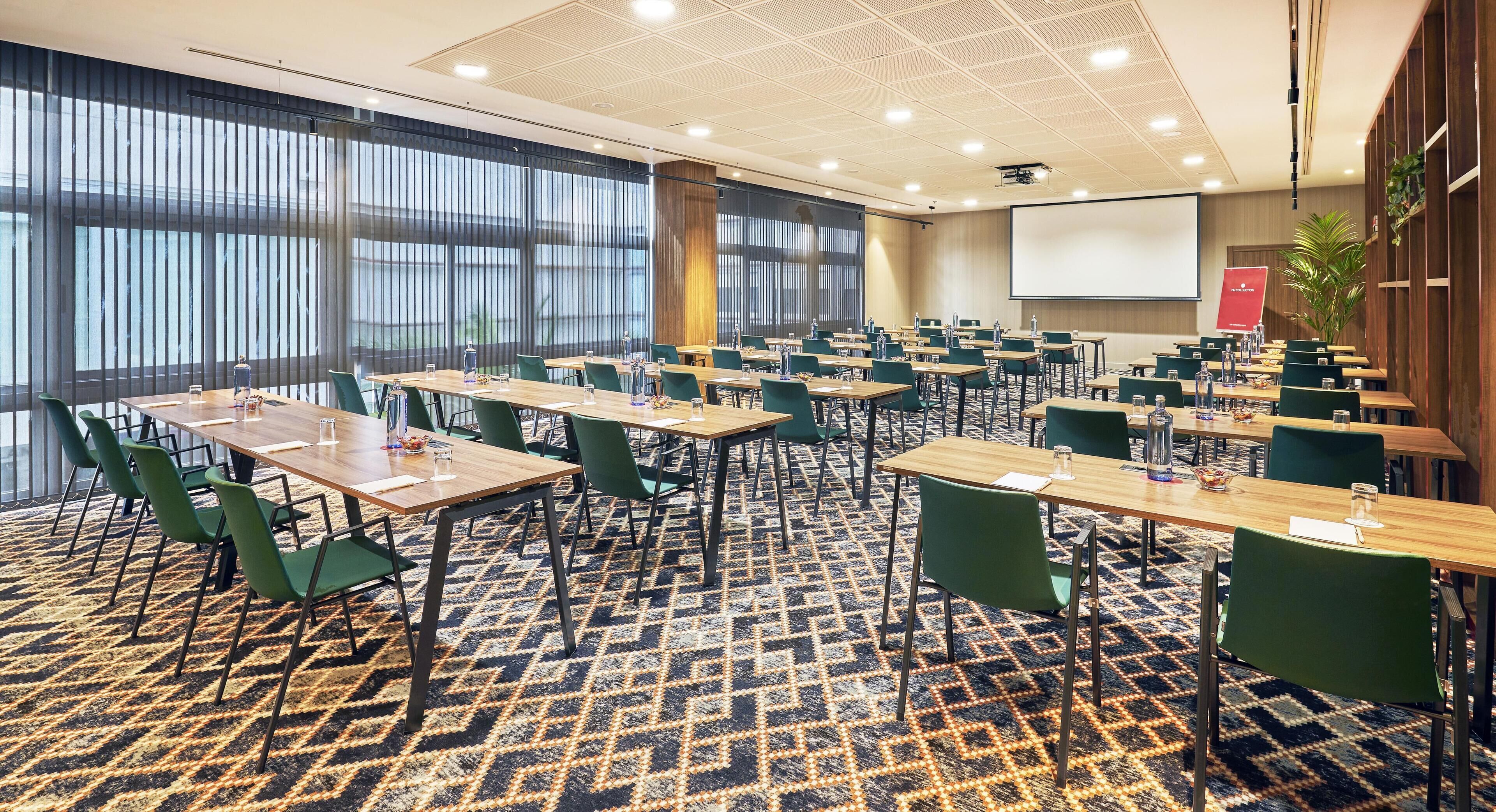 Uma sala de conferências com várias mesas e cadeiras, grandes janelas, uma tela de projetor e piso coberto de carpete.