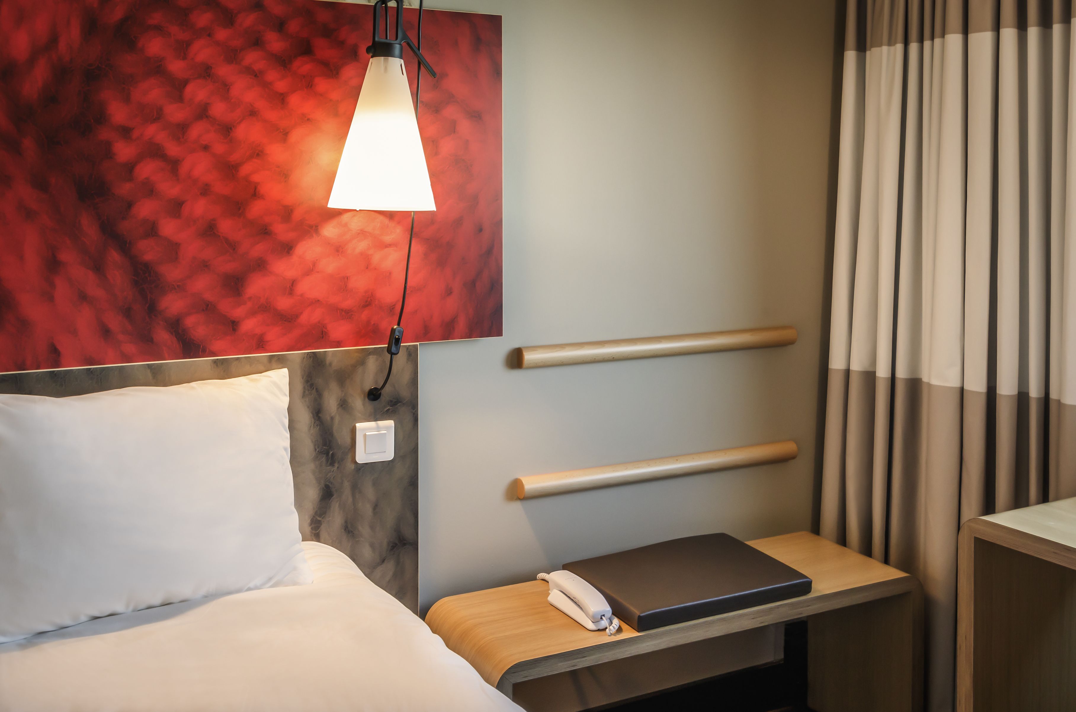 Quarto de hotel com cama, mesa de cabeceira, abajur e uma parede vermelha com textura.