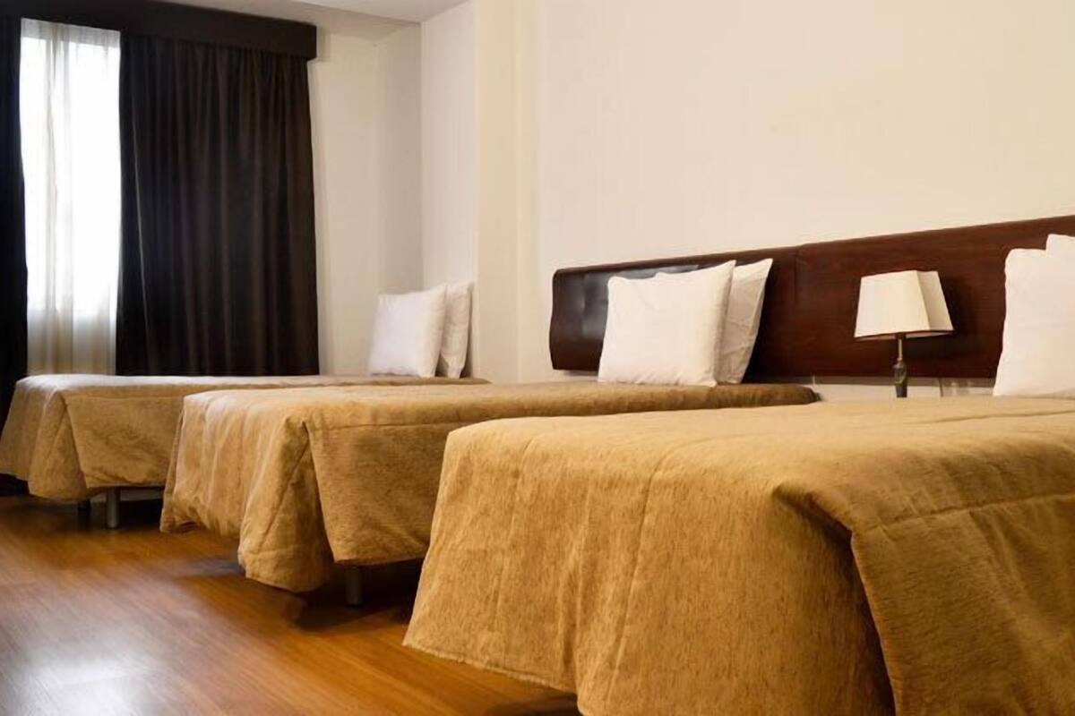 Quarto de hotel com três camas de solteiro, cada uma com colcha marrom e travesseiros brancos. Tem uma cabeceira de madeira no lado direito do quarto, e um abajur na mesa de cabeceira.
