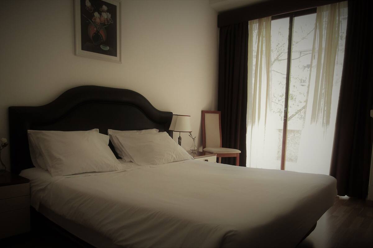 Uma cama bem arrumada com lençóis brancos, uma cabeceira escura e uma mesa de cabeceira com um abajur.