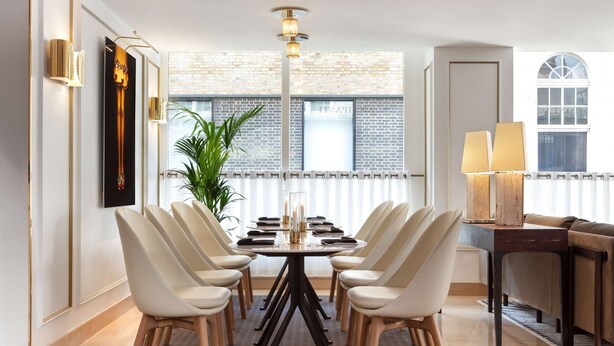Uma sala de jantar com uma mesa redonda, oito cadeiras, um vaso de planta e vista para o exterior por grandes janelas.