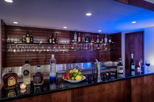 Uma área de bar com prateleiras de garrafas de bebidas, copos e uma cesta de frutas.
