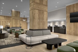 Um hall de hotel moderno com um sofá grande, pufes verdes, uma mesa de centro preta e uma mesa de recepção com uma TV de tela plana.