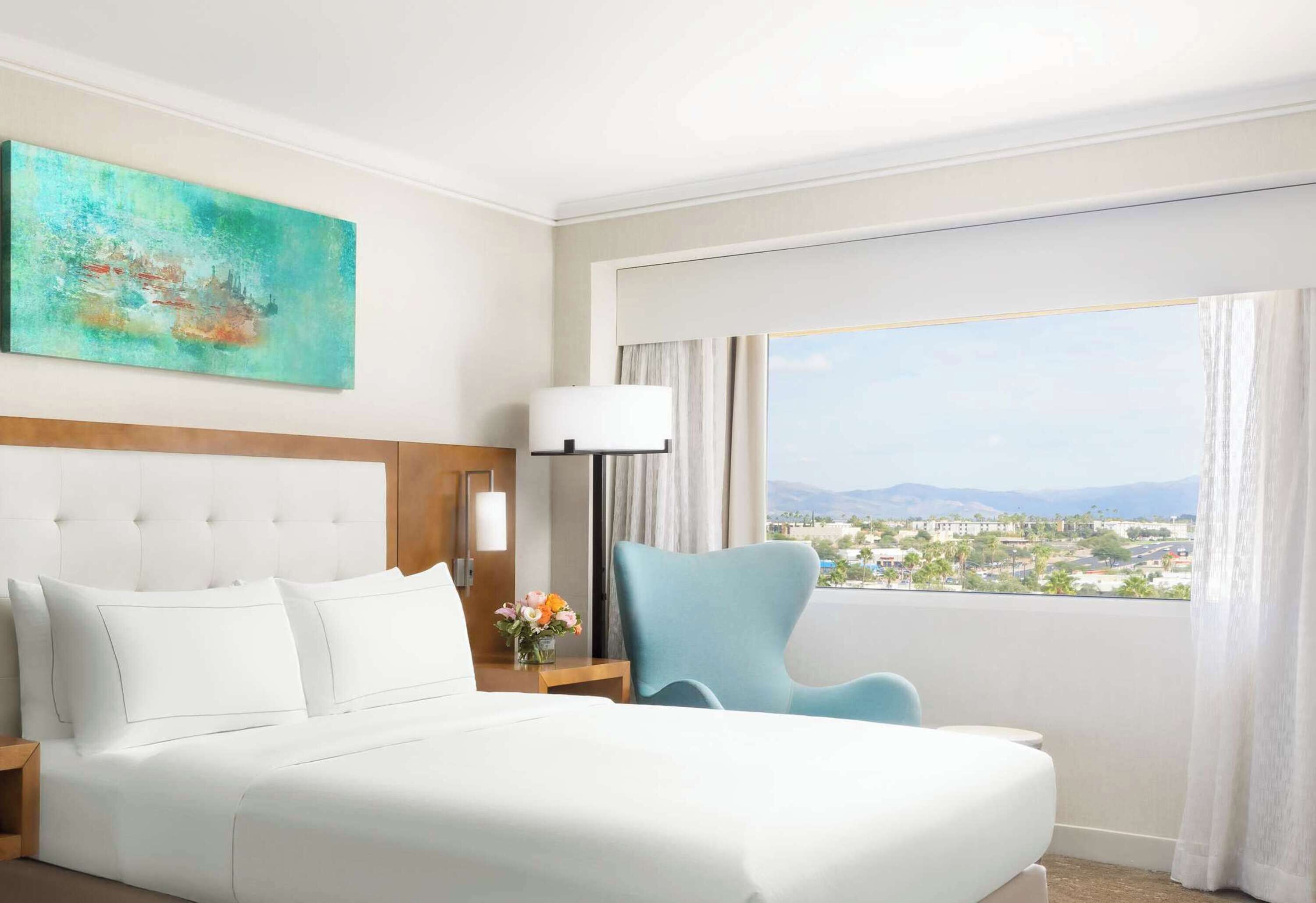 Quarto de hotel com uma cama, uma cadeira, um quadro, uma luminária e vista para a cidade.