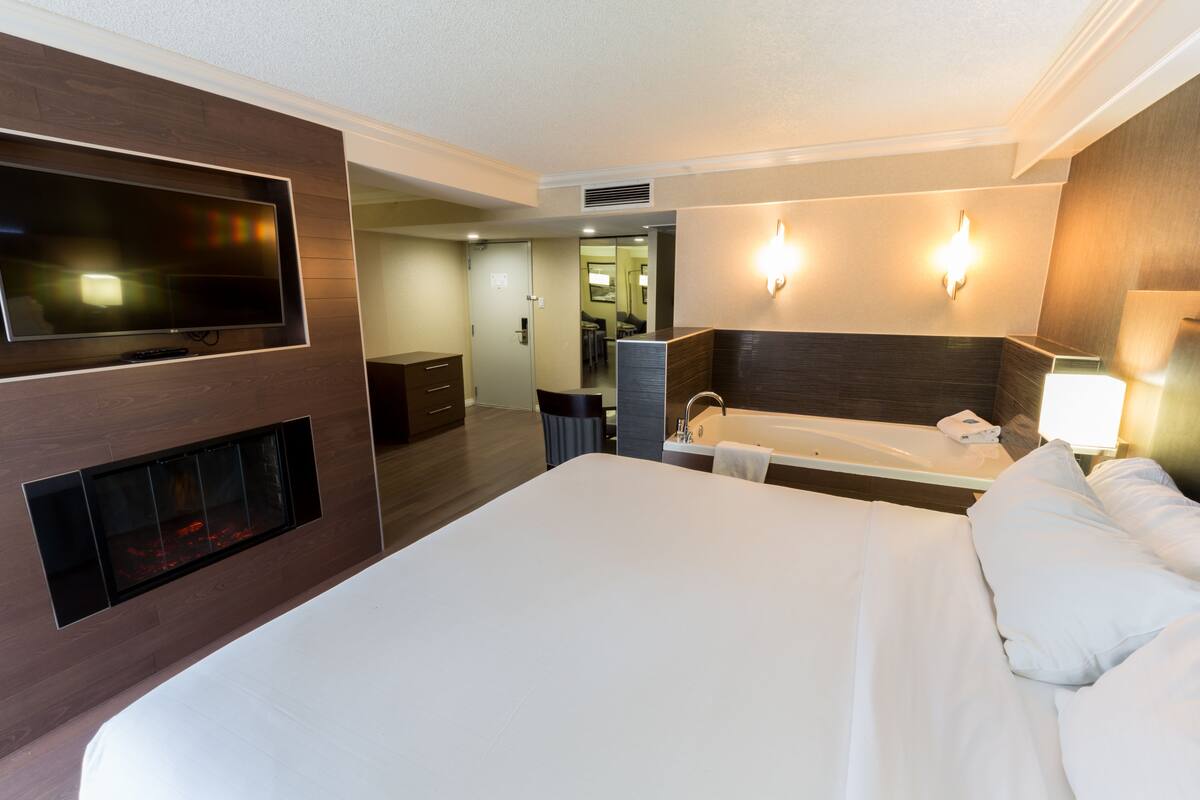 Quarto de hotel moderno com uma cama grande, televisão na parede, lareira embutida, banheiro com banheira e um closet espaçoso.