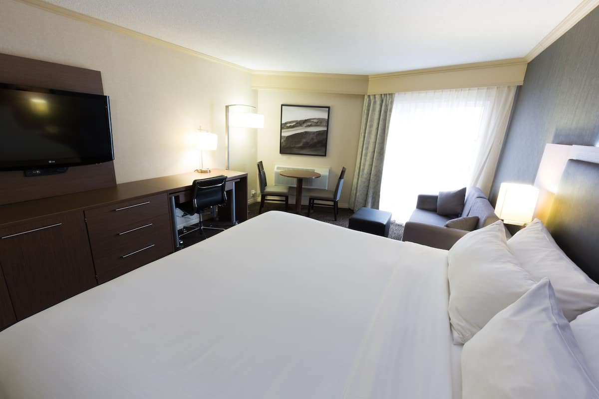 Quarto de hotel com uma cama grande, uma escrivaninha com cadeira, televisão e uma janela com cortinas.