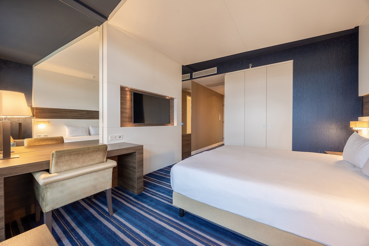 Quarto de hotel com uma cama grande, uma escrivaninha com cadeira, televisão e guarda-roupa.