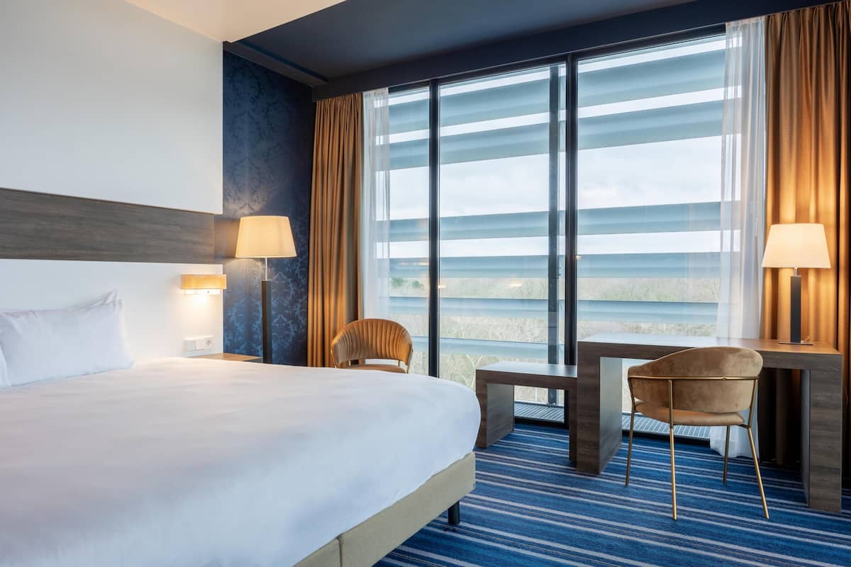 Quarto de hotel com uma cama grande, duas cadeiras, uma escrivaninha e vista através de amplas janelas.