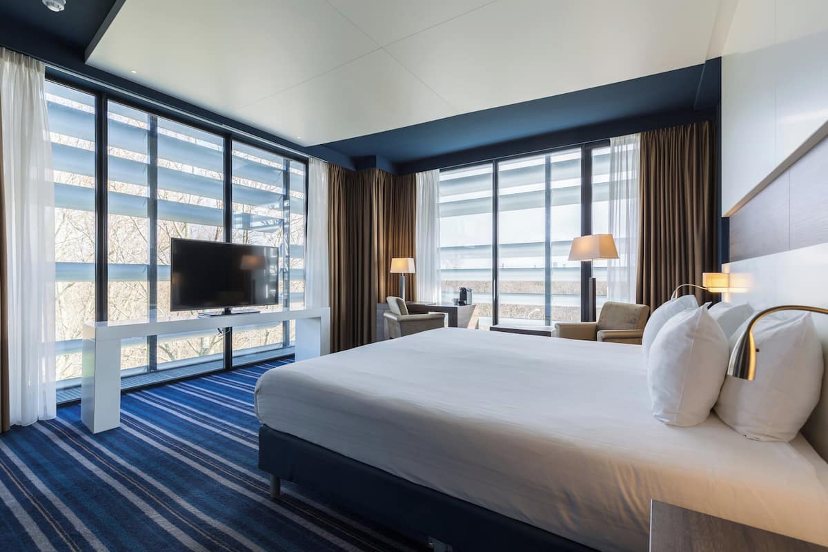 Quarto de hotel com uma cama grande, televisão de tela plana, escrivaninha com luminária e janelas amplas com cortinas.