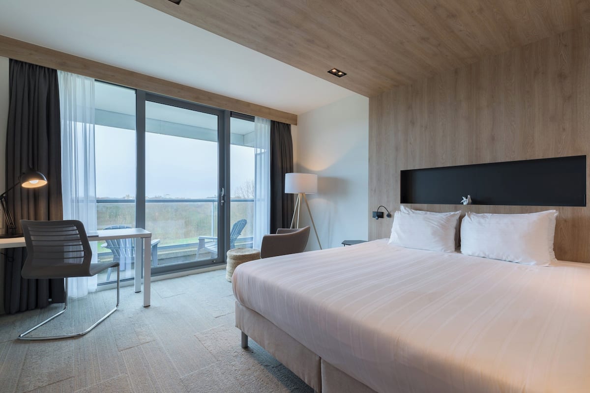 Quarto de hotel com uma cama grande, uma escrivaninha com cadeira, uma luminária e vista pela janela.