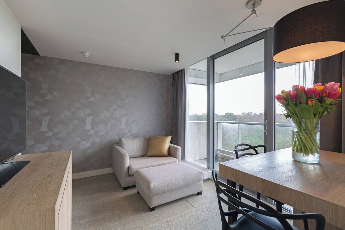 Uma sala de estar moderna com um sofá, um banquinho, uma mesa de jantar e cadeiras. Há uma televisão em um suporte de madeira, um vaso com flores na mesa e uma grande janela com vista.