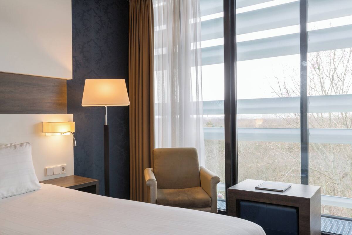Quarto de hotel com cama, uma cadeira, mesa de cabeceira com abajur e vista para árvores por grandes janelas.
