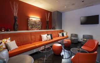 Uma área de estar moderna com um sofá laranja comprido, diversas opções de assento incluindo mesas redondas e cadeiras, uma TV de tela plana na parede e vasos decorativos em uma estante.