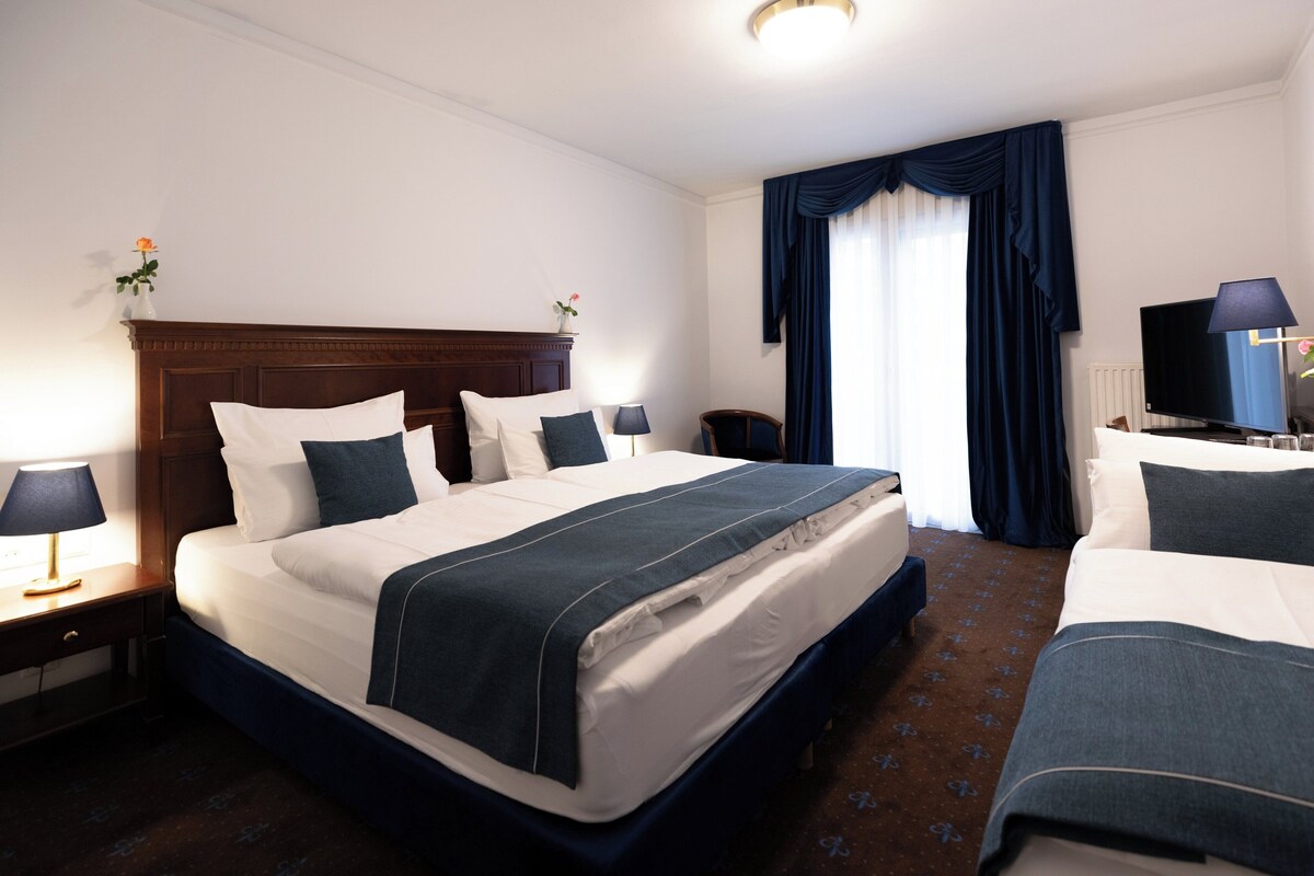 Quarto de hotel com uma cama grande, uma cama menor, uma televisão e uma janela com cortinas.