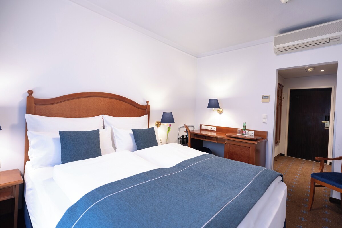 Quarto de hotel com uma cama grande, uma mesa de cabeceira com abajur, uma escrivaninha e uma cadeira.