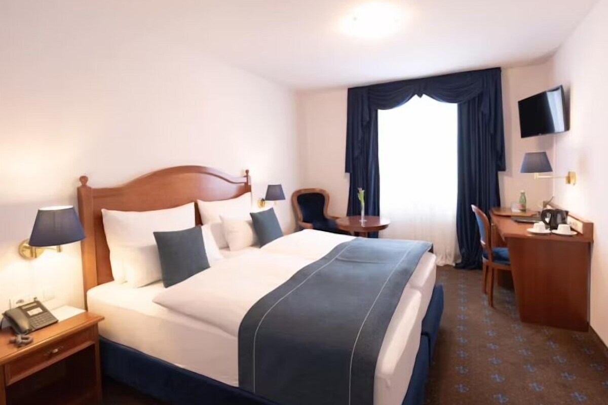 Quarto de hotel com uma cama grande, mesas de cabeceira, escrivaninha, cadeira, televisão e janela com cortinas.