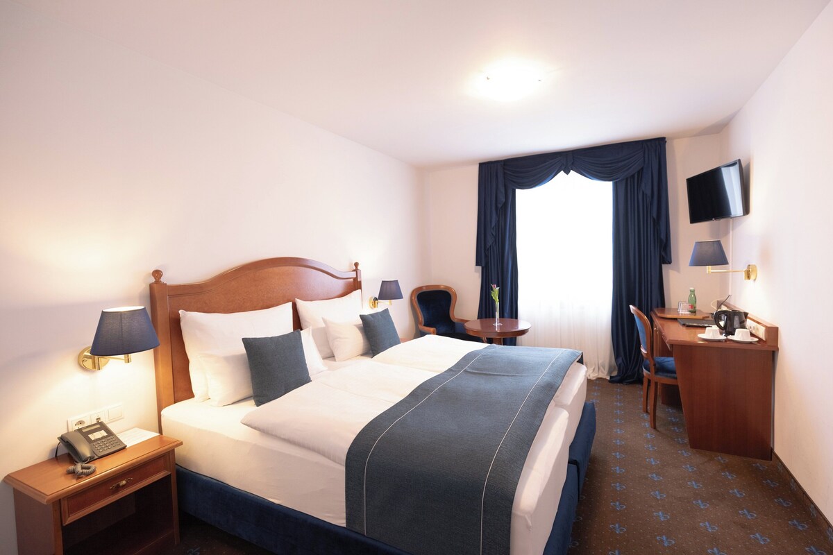 Quarto de hotel com uma cama grande, mesas de cabeceira, escrivaninha, uma cadeira, televisão e uma janela com cortinas.