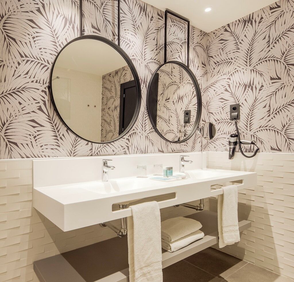 Casa de banho com uma grande pia branca, dois espelhos redondos e um secador de cabelo fixado na parede.