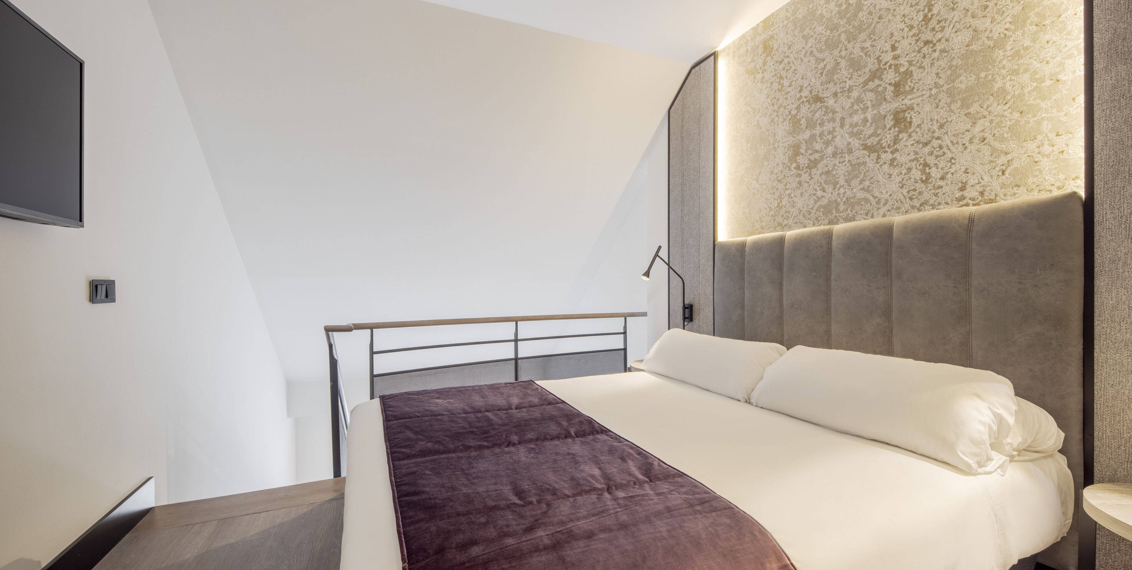 Quarto de hotel moderno com cama, cabeceira com parede texturizada, TV de tela plana na parede e uma pequena mesa de cabeceira.
