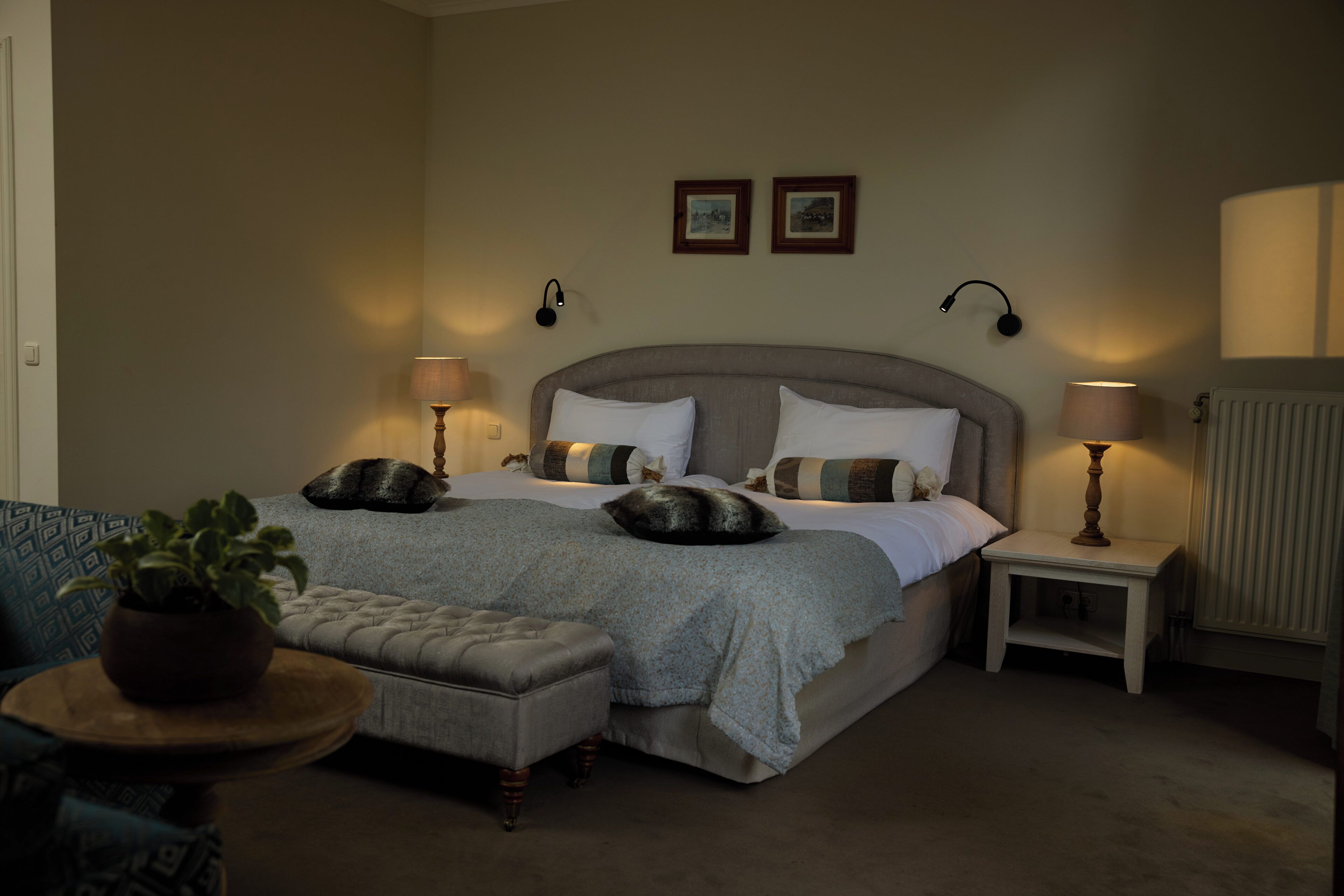 Quarto com uma cama grande, duas luminárias de cabeceira, uma mesinha com uma planta e um radiador.
