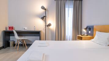 Quarto de hotel com cama, escrivaninha, cadeira, luminária e janela com cortinas.