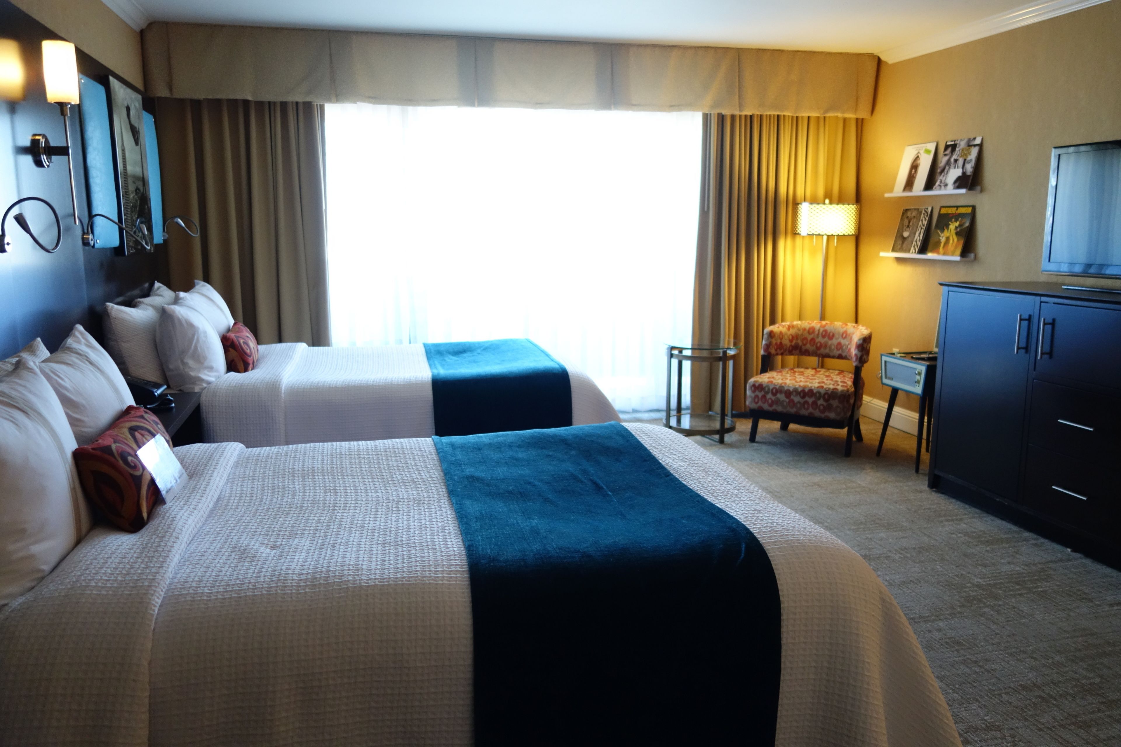 Quarto de hotel com duas camas, uma escrivaninha, uma cadeira, uma televisão e uma janela com cortinas.