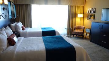 Quarto de hotel com duas camas, uma escrivaninha, uma cadeira, uma televisão e uma janela com cortinas.