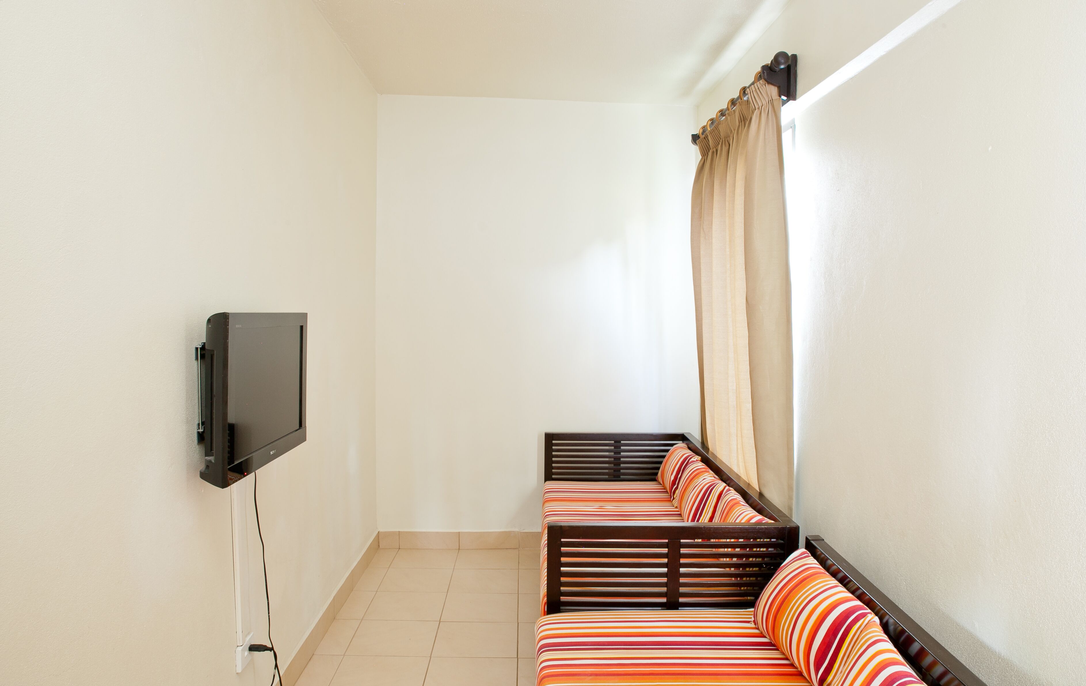 Um quarto pequeno e bem iluminado, com televisão na parede, sofá com almofadas listradas e cortina na janela.