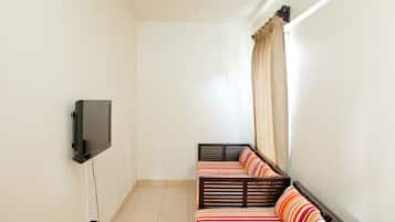 Um quarto pequeno e bem iluminado, com televisão na parede, sofá com almofadas listradas e cortina na janela.