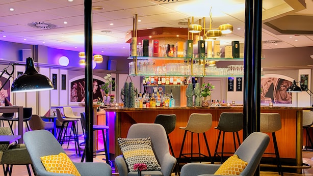 Uma área de bar com diversas opções de assento, balcão e variedade de garrafas e copos nas prateleiras.
