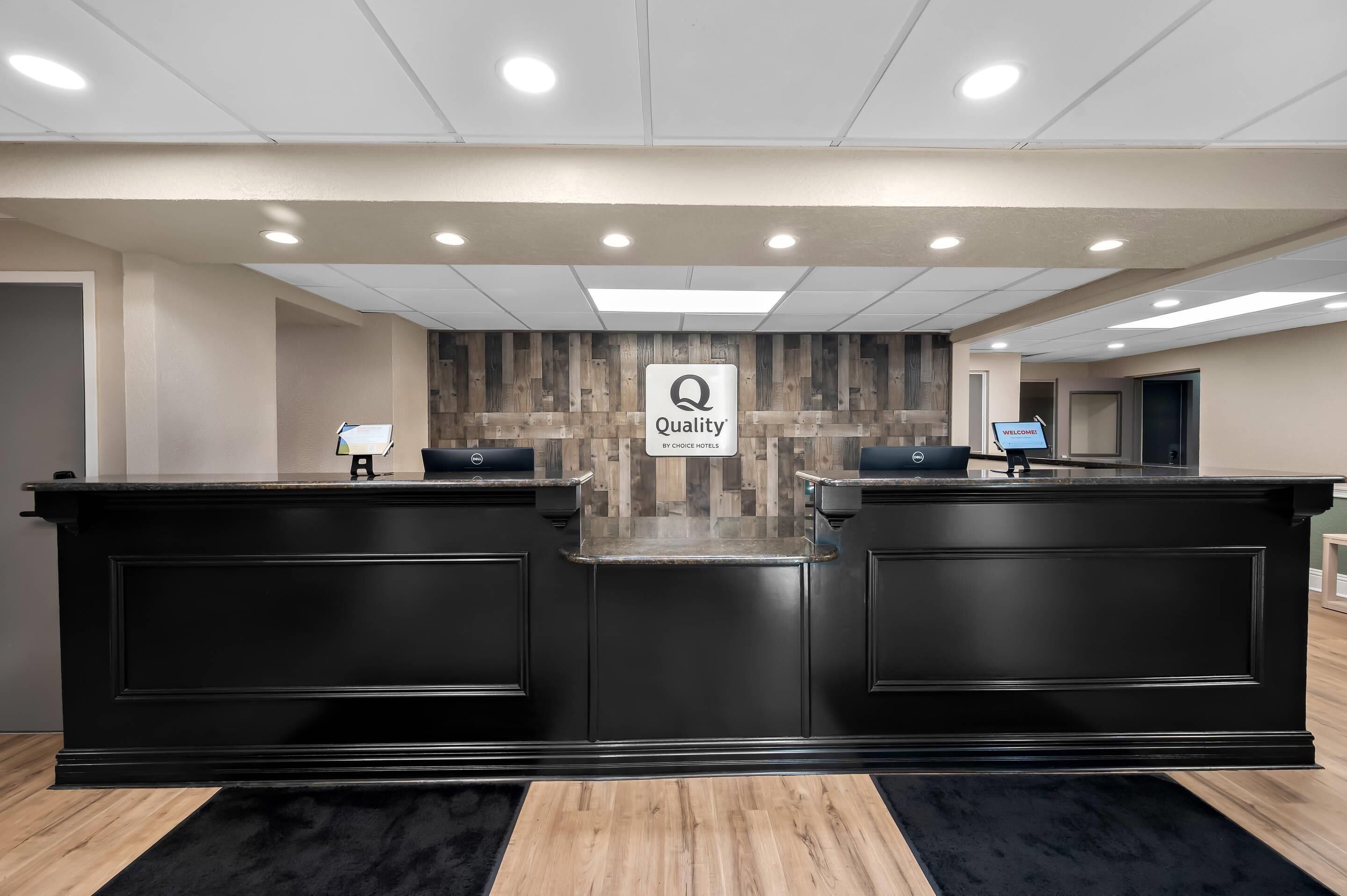 Uma área de recepção de hotel com dois balcões, uma placa com a inscrição 'Quality by Choice Hotels' e um letreiro na parede com a palavra 'BEM-VINDOS!'.