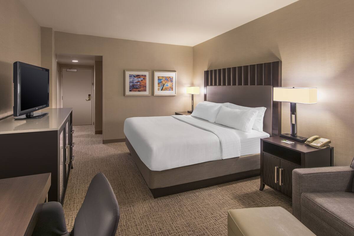 Quarto de hotel com cama, escrivaninha com televisão, cadeira, sofá e mesa de cabeceira com abajur e telefone.