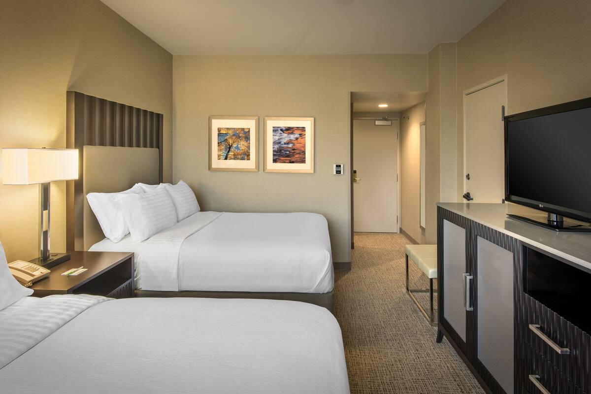 Quarto de hotel com duas camas, televisão, escrivaninha com abajur e duas quadros na parede.