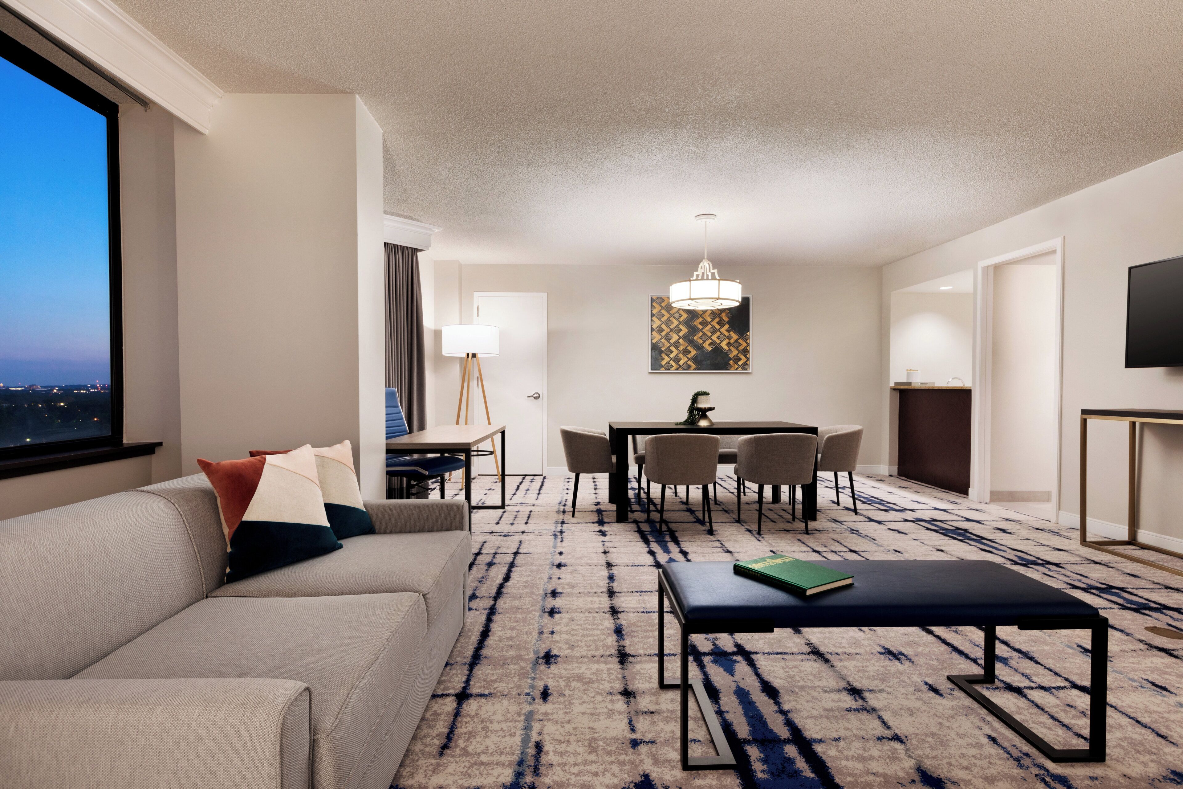Quarto de hotel moderno com sofá, mesa de jantar, cadeiras, mesa de centro, TV e uma grande janela com vista para a cidade.