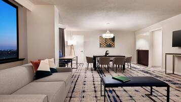 Quarto de hotel moderno com sofá, mesa de jantar, cadeiras, mesa de centro, TV e uma grande janela com vista para a cidade.